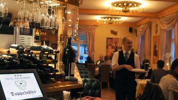Interior de restaurante de hotel com área de bar, garrafas de vinho e um garçom atendendo os hóspedes.