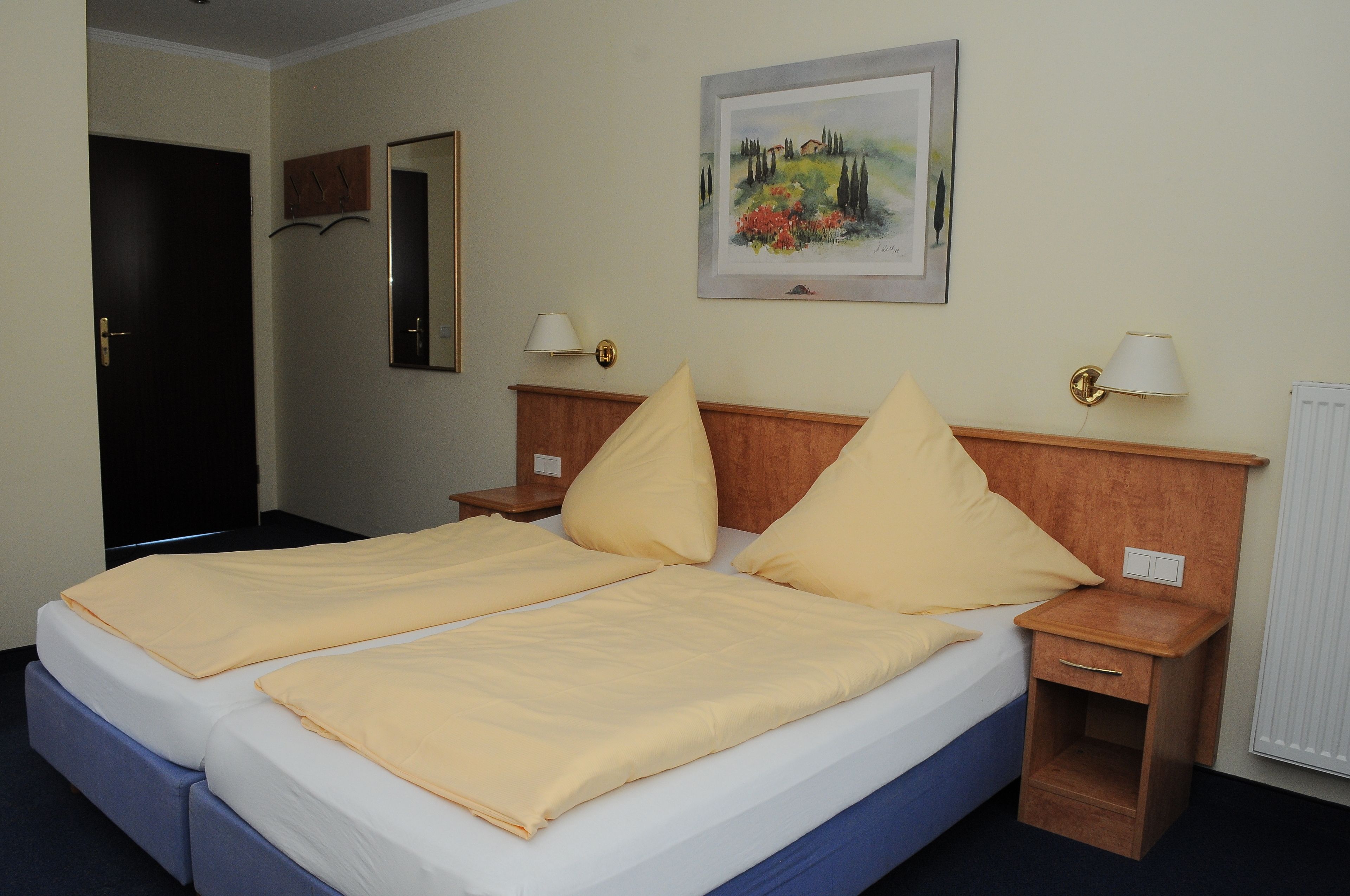 Uma cama de casal com duas almofadas, uma mesinha de cabeceira de madeira, uma mesa lateral com abajur e um quadro emoldurado na parede.