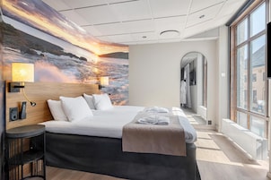 Quarto de hotel com uma cama grande, vista da cidade através de amplas janelas, um mural com um pôr do sol sobre a água na parede e uma pequena mesa lateral com uma luminária.