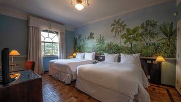 Quarto de hotel com duas camas, uma escrivaninha com abajur, uma janela com cortinas e um mural de plantas tropicais na parede.