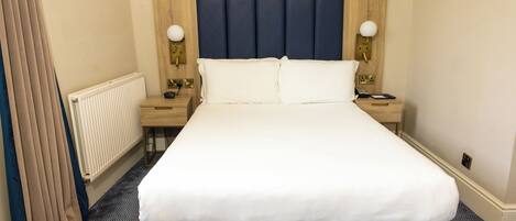 Quarto de hotel com uma cama grande, cabeceira de madeira, duas mesas de cabeceira com abajures, um radiador e uma janela com cortinas.