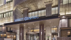 Uma entrada de hotel moderna com fachada curva, um destaque para a placa "Hyatt Centric" e um conjunto de degraus que levam à entrada.