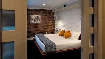 Quarto com parede de tijolos, cama com roupa de cama branca e almofadas laranja, manta cinza e luminária pendente.