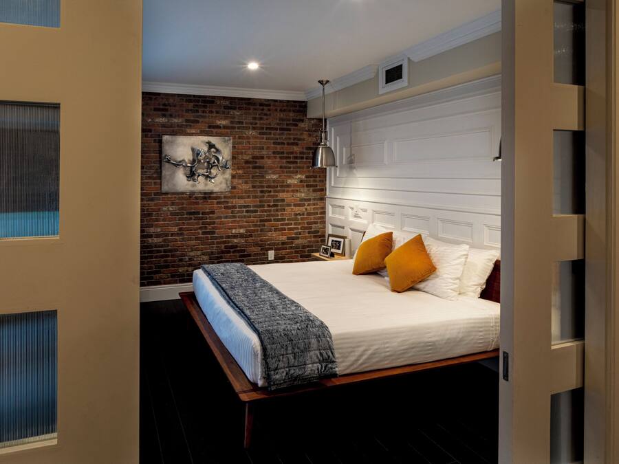 Quarto com parede de tijolos, cama com roupa de cama branca e almofadas laranja, manta cinza e luminária pendente.