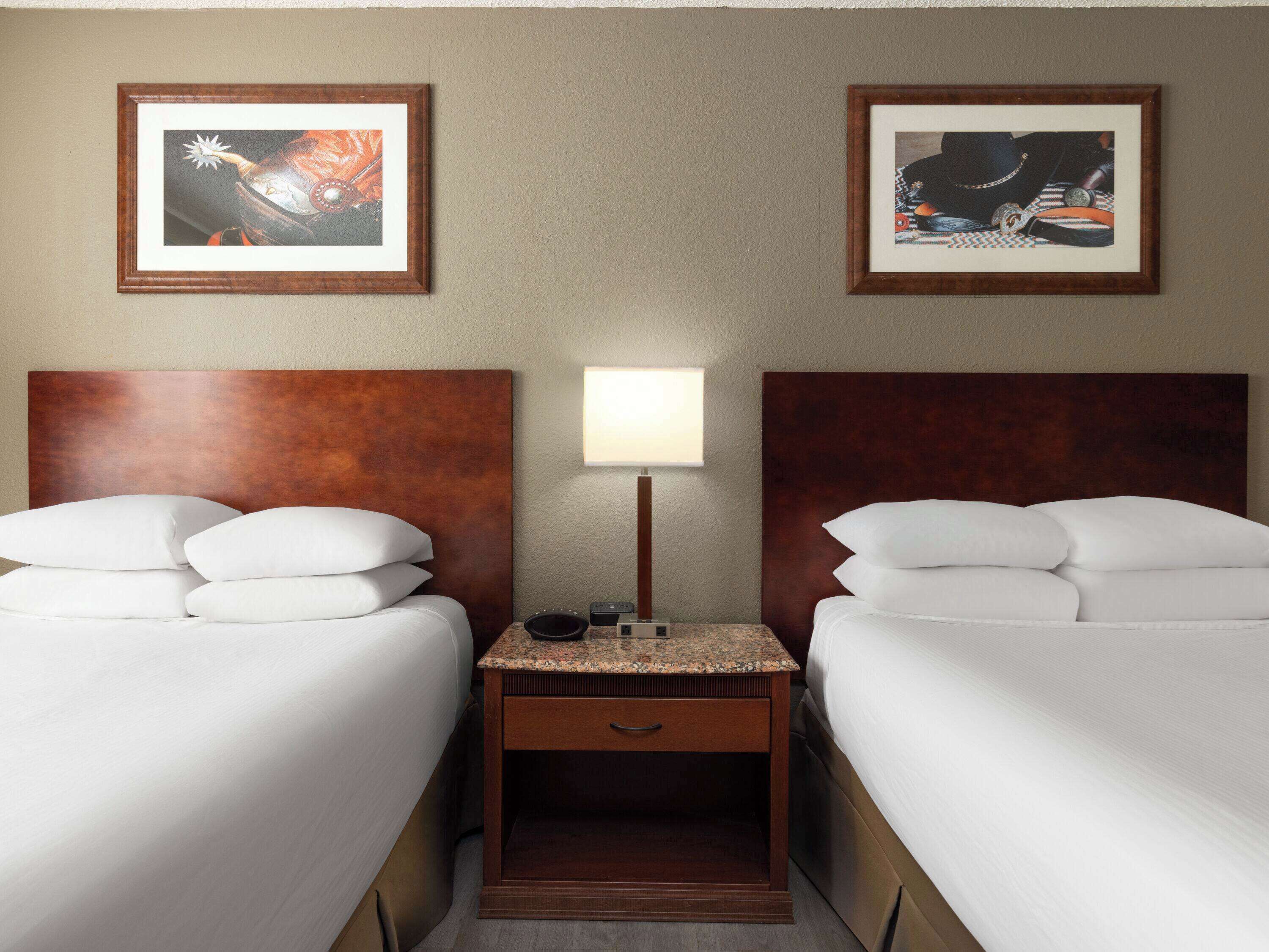 Quarto de hotel com duas camas, uma mesa de cabeceira com abajur e duas fotografias emolduradas na parede.