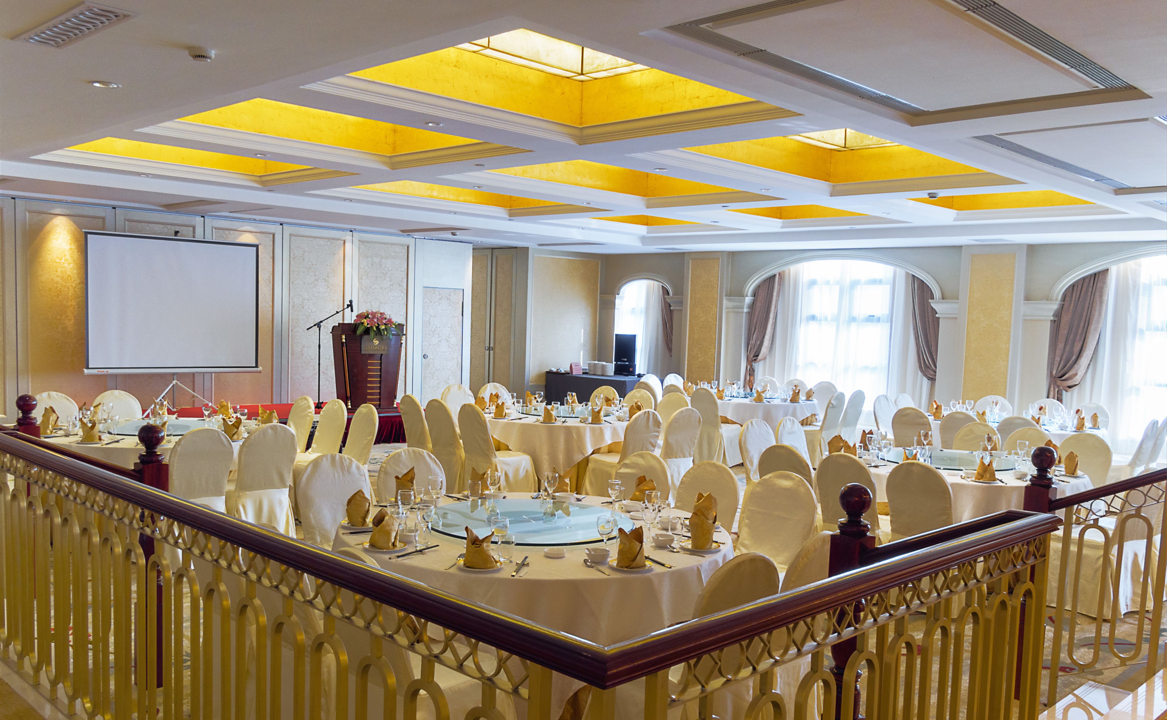 Uma sala de banquetes com mesas redondas, cadeiras brancas e um palco com um pódio.