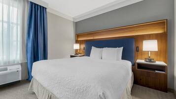 Quarto de hotel com uma cama grande, cabeceira de madeira, duas mesas de cabeceira com abajures, uma janela com cortinas azuis e um ar-condicionado fixado na parede.