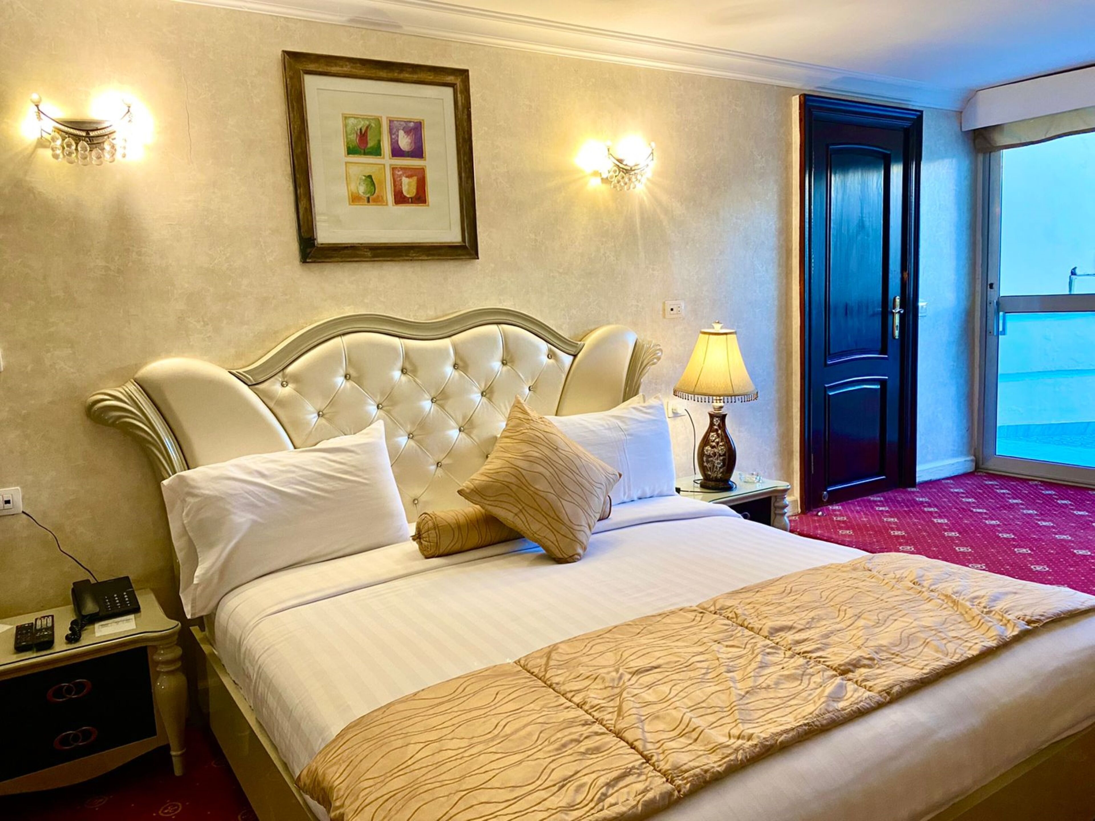 Quarto de hotel com uma cama grande, mesa de cabeceira com abajur, quadro na parede e porta que dá para uma varanda.