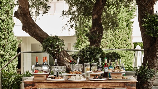 Mesa de jantar ao ar livre posta com diversos alimentos, garrafas e copos, cercada por árvores e um edifício ao fundo.
