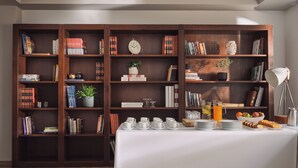 Quarto com uma grande estante de madeira repleta de livros e objetos decorativos, uma mesa com toalha branca e xícaras de chá, e um abajur à direita.