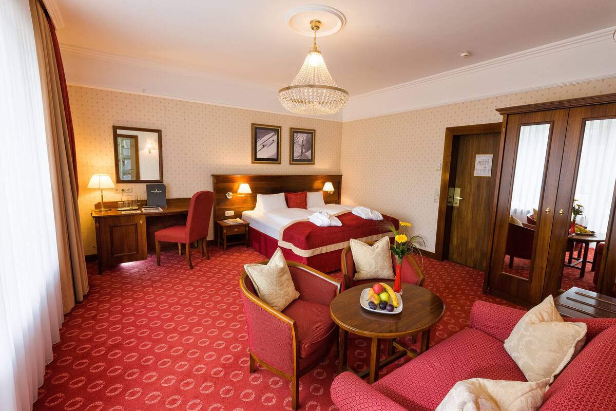 Quarto de hotel com cama, escrivaninha, poltrona, sofá, mesa com frutas e um candelabro.