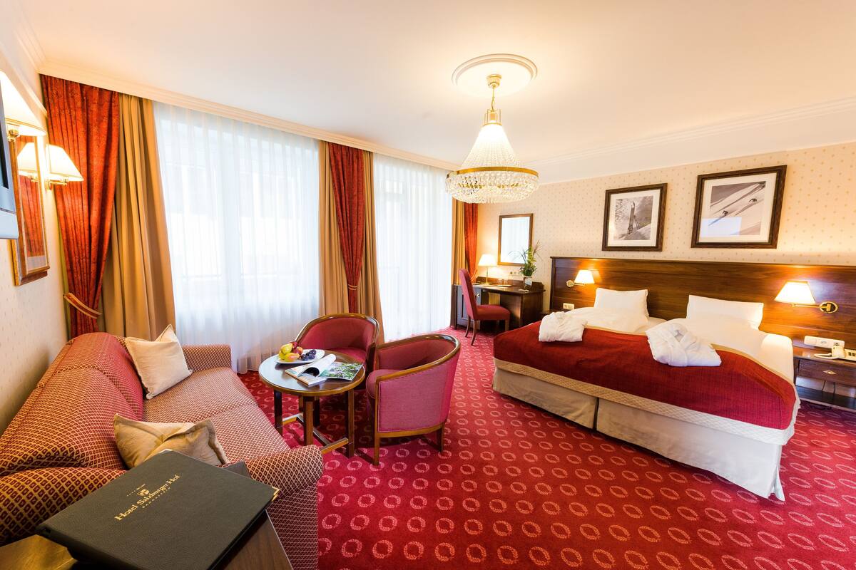 Quarto de hotel com carpete vermelho, cama com roupa de cama branca, um sofá, uma mesa pequena com uma revista e uma cadeira.