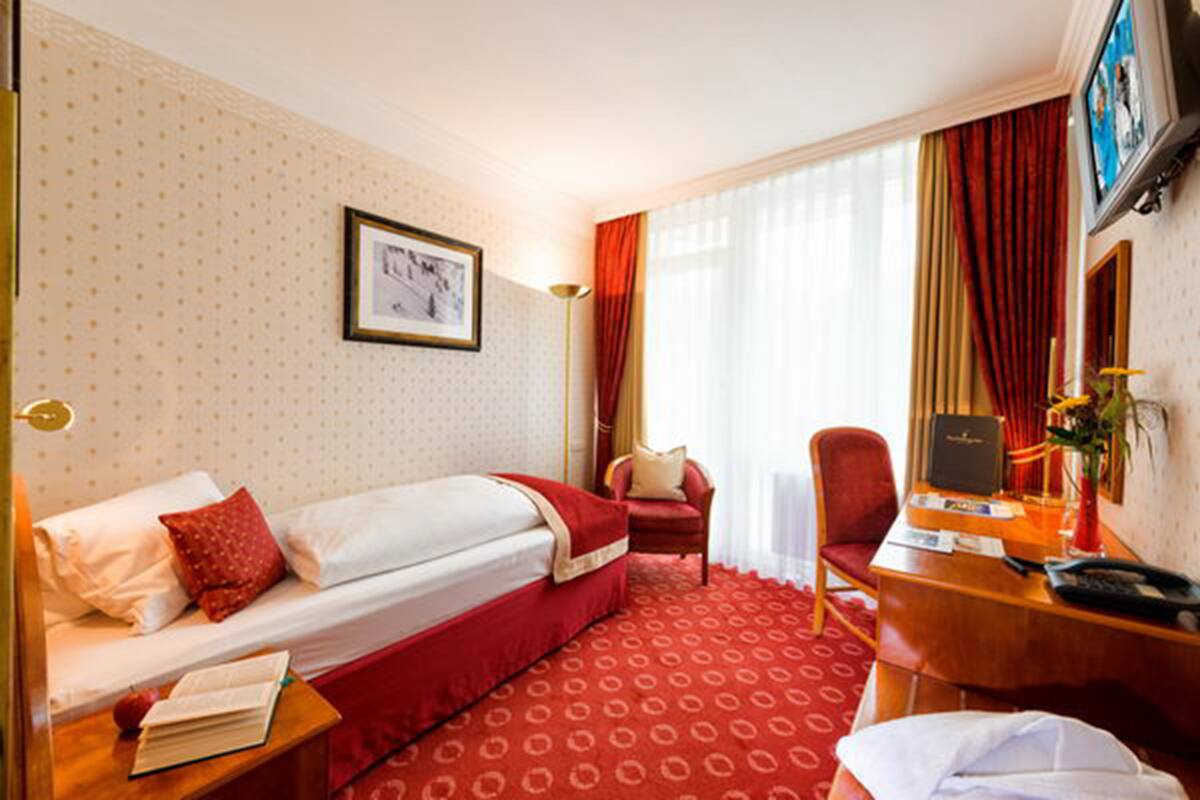 Quarto de hotel com cama, escrivaninha, cadeira e uma pequena mesa com um livro e um vaso de flores.