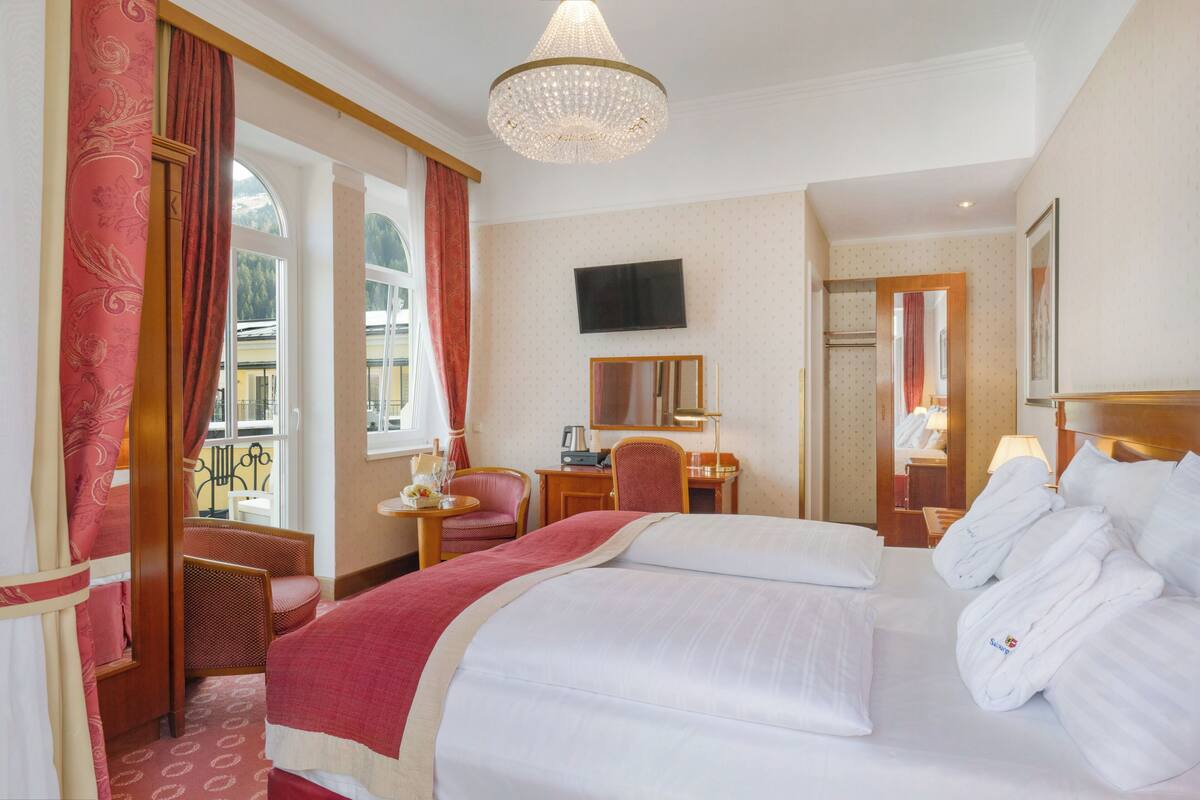 Quarto de hotel com cama, secretária, cadeira, televisão e lustre.