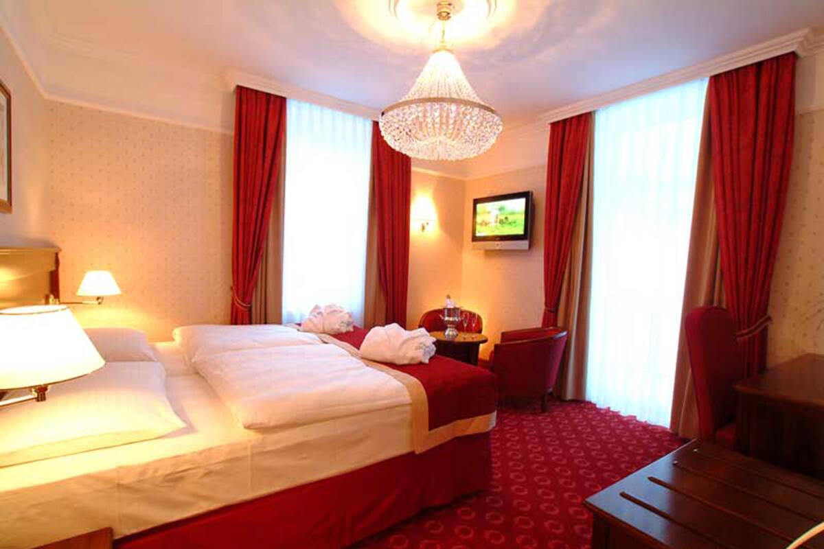 Quarto de hotel com uma cama grande, uma escrivaninha, uma cadeira, uma televisão e um candelabro.
