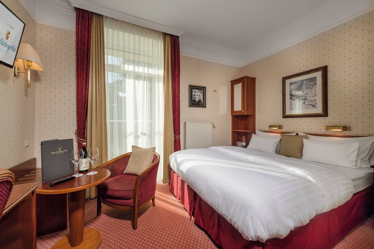 Quarto de hotel com cama, escrivaninha com laptop, cadeira, mesa de cabeceira, luminária e janela com cortinas.