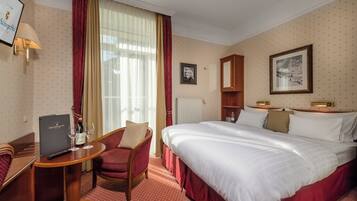 Quarto de hotel com cama, escrivaninha com laptop, cadeira, mesa de cabeceira, luminária e janela com cortinas.