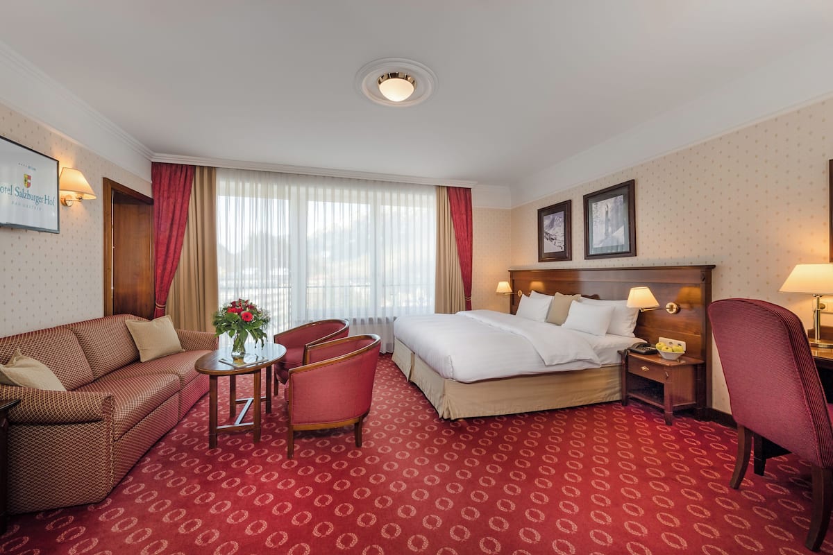 Quarto de hotel com cama, sofá, poltrona, mesa pequena com um vaso de flores, mesa de cabeceira com abajur e janela com cortinas.