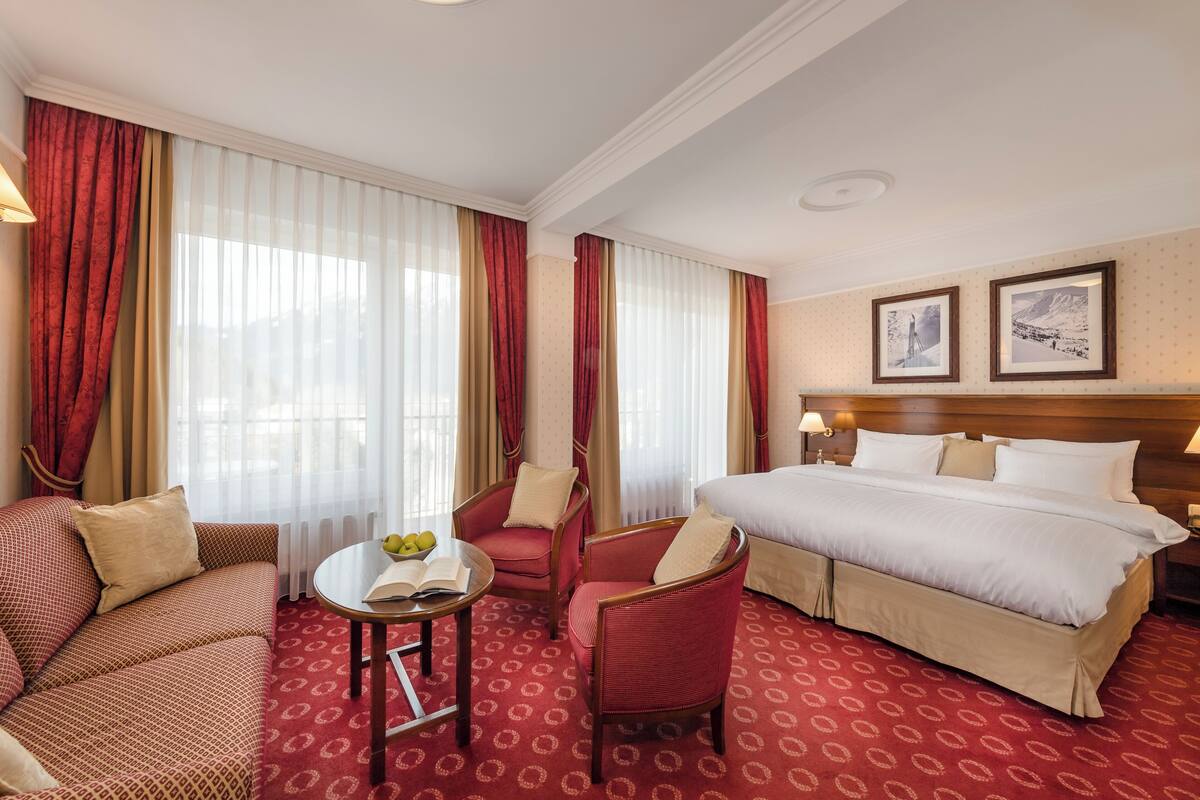Quarto de hotel com cama, duas cadeiras, uma mesinha com um livro e frutas, e uma janela grande com cortinas.