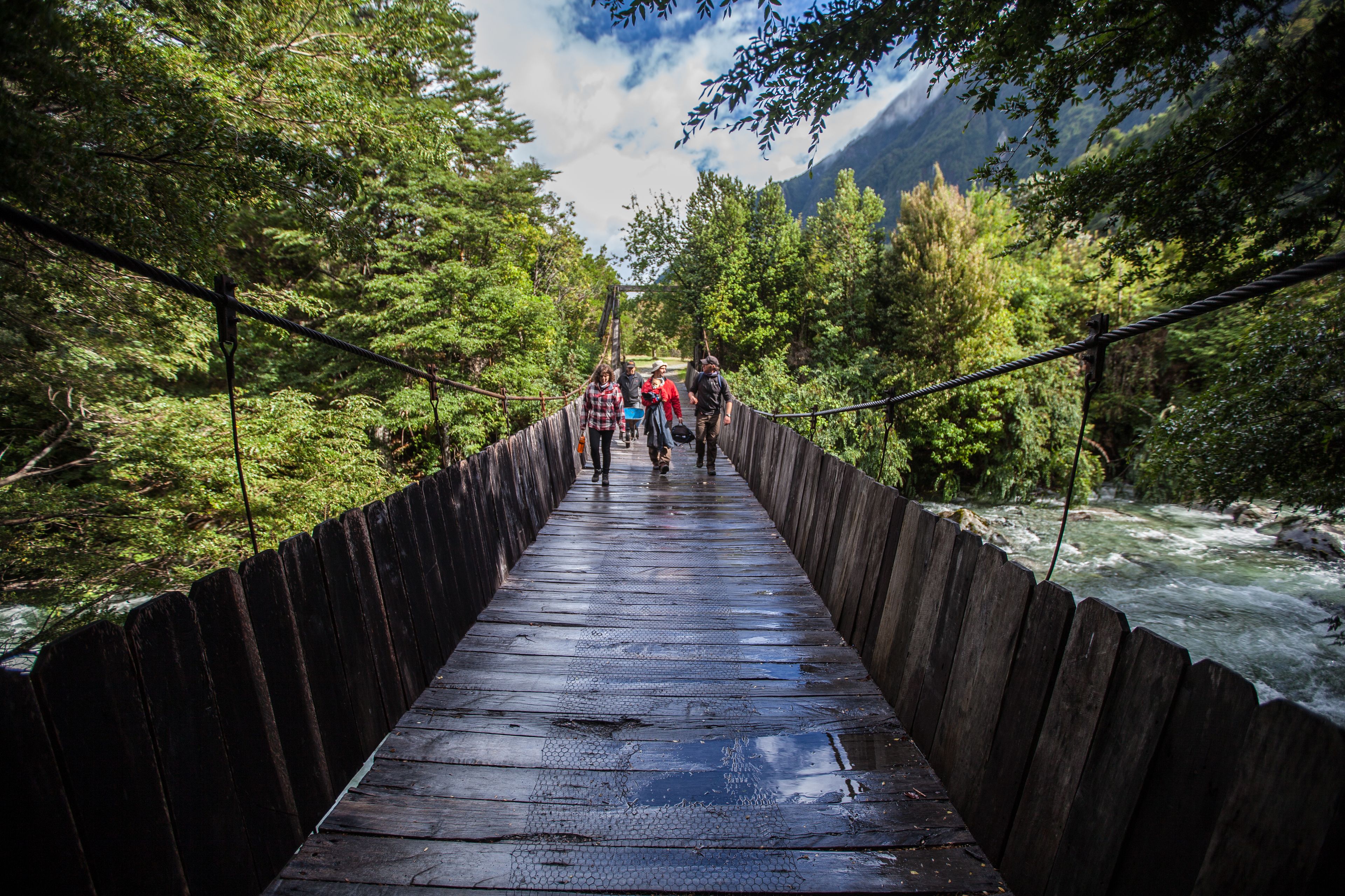 Uma ponte suspensa de madeira sobre um rio, rodeado por uma floresta densa e montanhas.