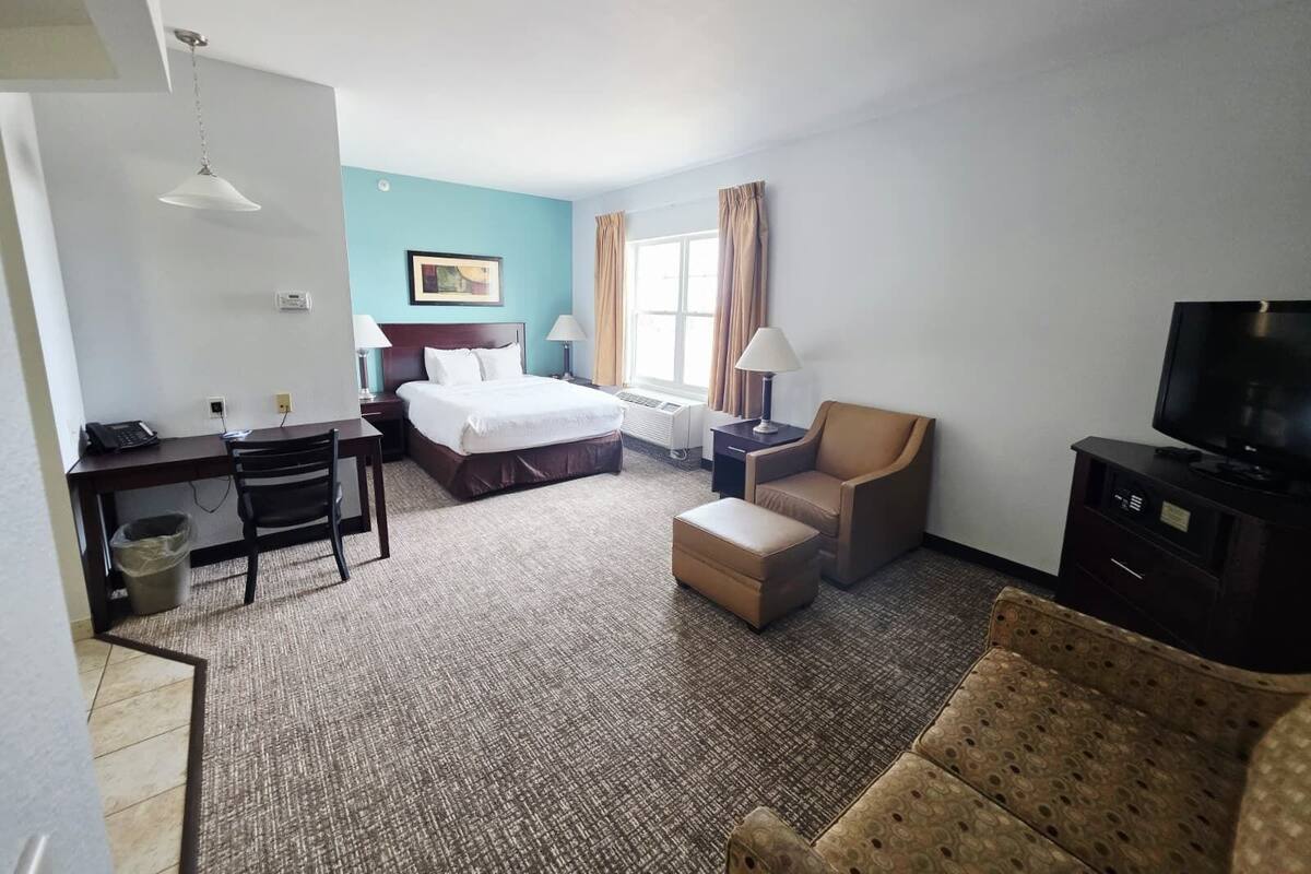 Quarto de hotel com cama, escrivaninha, cadeira, banquinho, televisão e janela com cortinas.