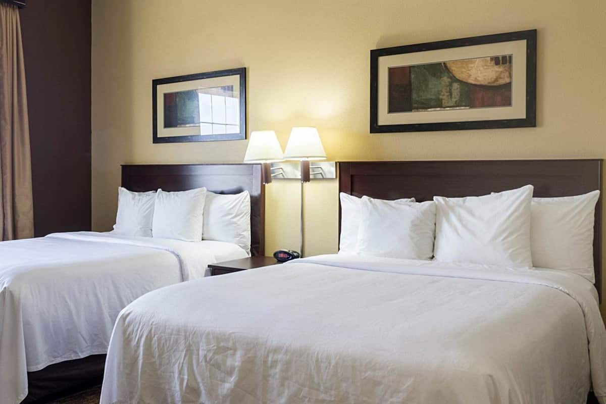 Quarto de hotel com duas camas, uma mesa de cabeceira com abajur e duas fotos emolduradas na parede.