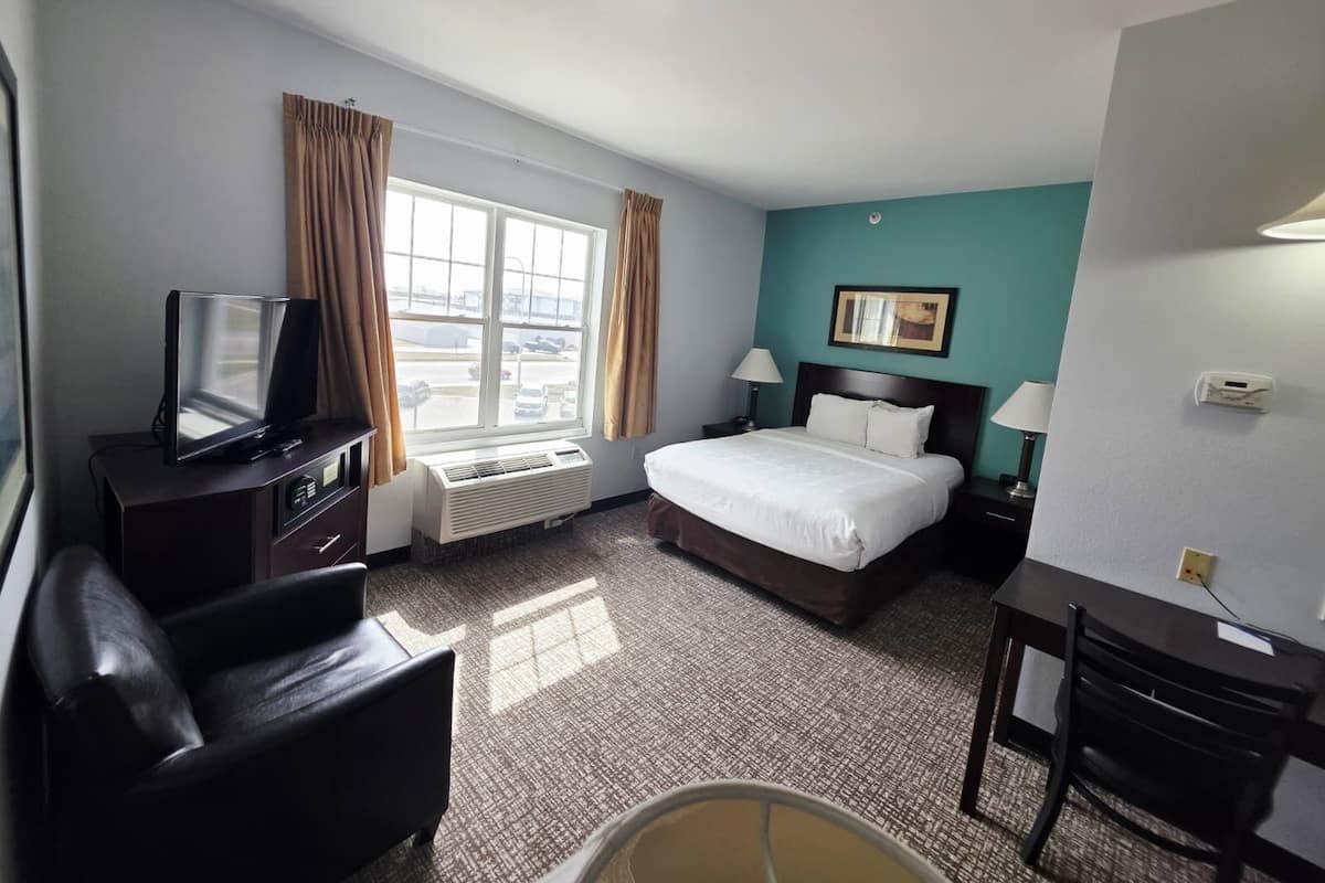 Quarto de hotel com cama, escrivaninha, cadeira, televisão e janela com cortinas.
