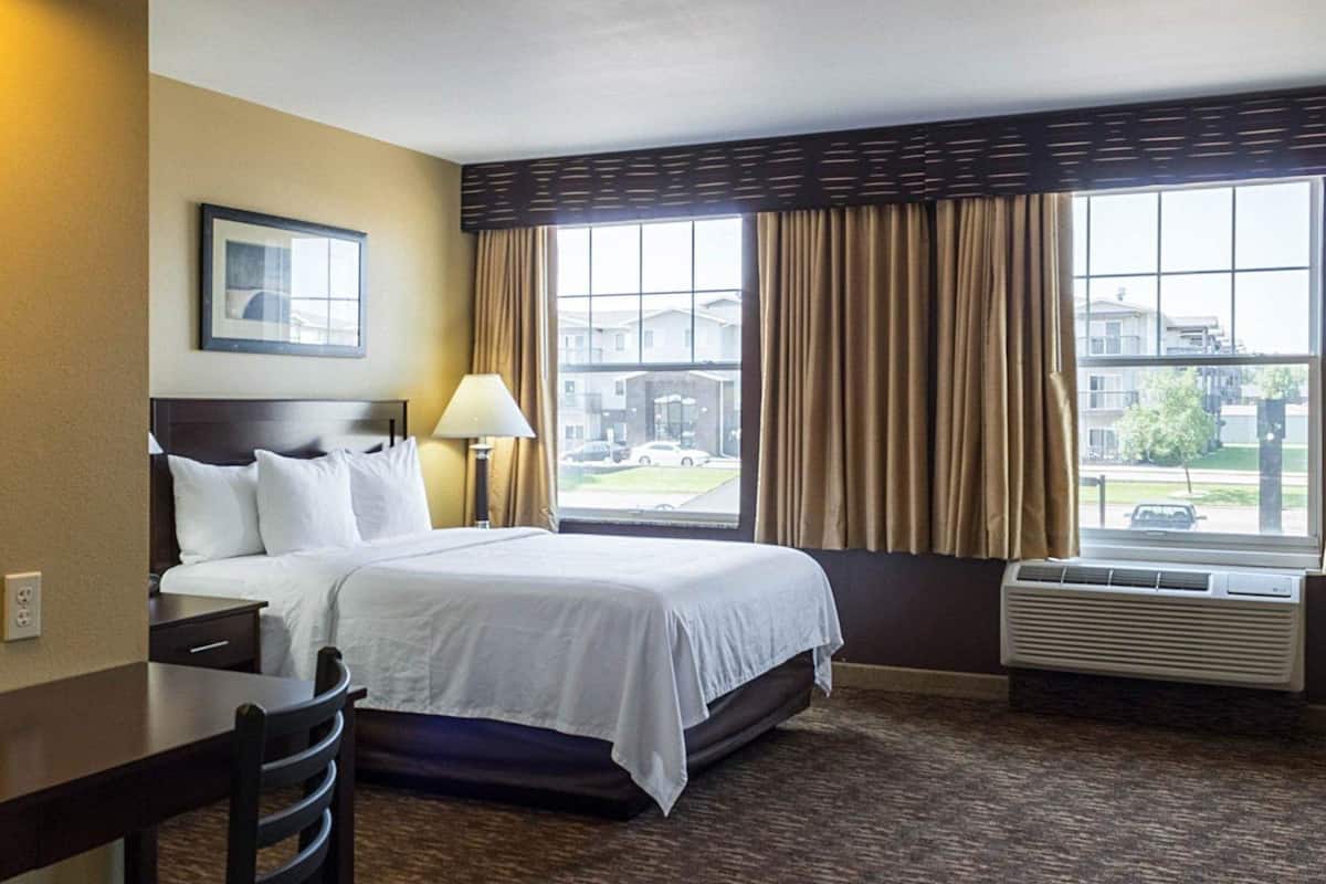 Quarto de hotel com cama, escrivaninha, cadeira, janela com cortinas e ar condicionado.