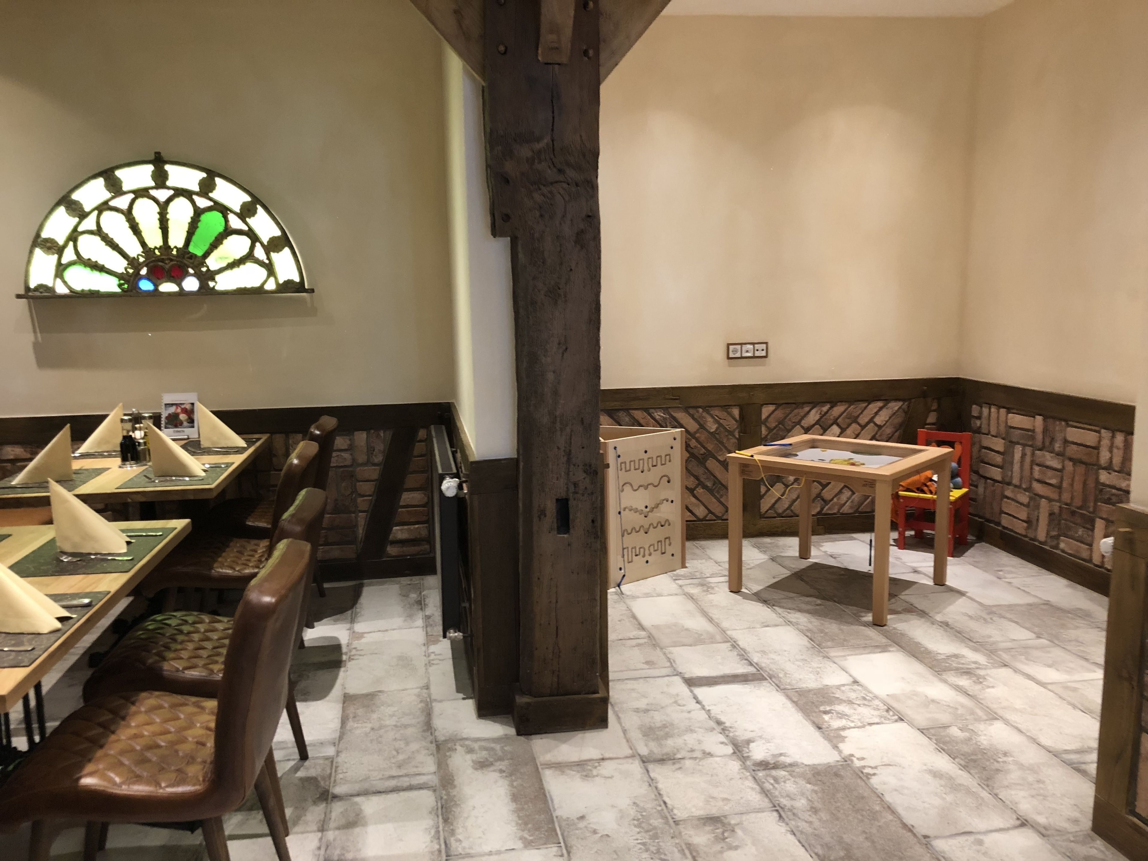 Interior de restaurante com mesas e cadeiras de madeira, uma janela de vitrais e uma parede de pedra.
