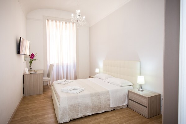 Quarto de hotel moderno com uma cama grande, mesas de cabeceira, escrivaninha, uma cadeira e uma TV na parede.