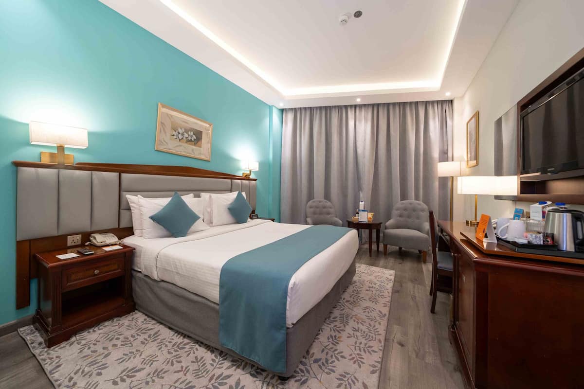 Quarto de hotel com cama, abajures, escrivaninha com cafeteira, televisão e uma cadeira.