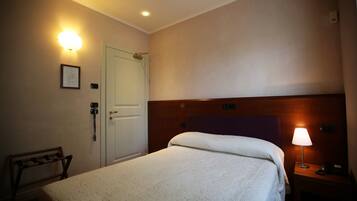 Quarto de hotel com cama, mesa de cabeceira com luminária, cabeceira de madeira, porta e luminária fixada na parede.
