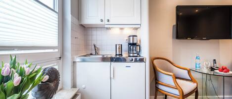 Uma cozinha compacta com armários brancos, uma máquina de café embutida e uma mesa de vidro com uma cadeira.