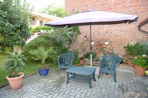 Um pátio externo com mesa, duas cadeiras, um guarda-sol roxo e plantas em vasos.