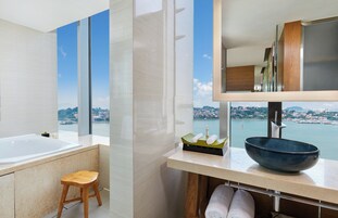 Uma casa de banho moderna com uma banheira, um banquinho de madeira, uma janela ampla com vista para a paisagem urbana e uma pia com uma bacia.
