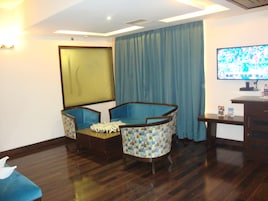 Quarto com chão de madeira, televisão na parede e duas cadeiras azuis com uma mesa estampada no centro.