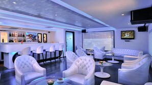 Uma área de estar moderna com sofás e poltronas de couro branco, um bar com garrafas de bebidas e uma televisão fixada na parede.
