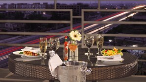 Mesa de jantar posta com vários copos, uma garrafa e um arranjo floral, com vista para um horizonte urbano onde se veem semáforos.