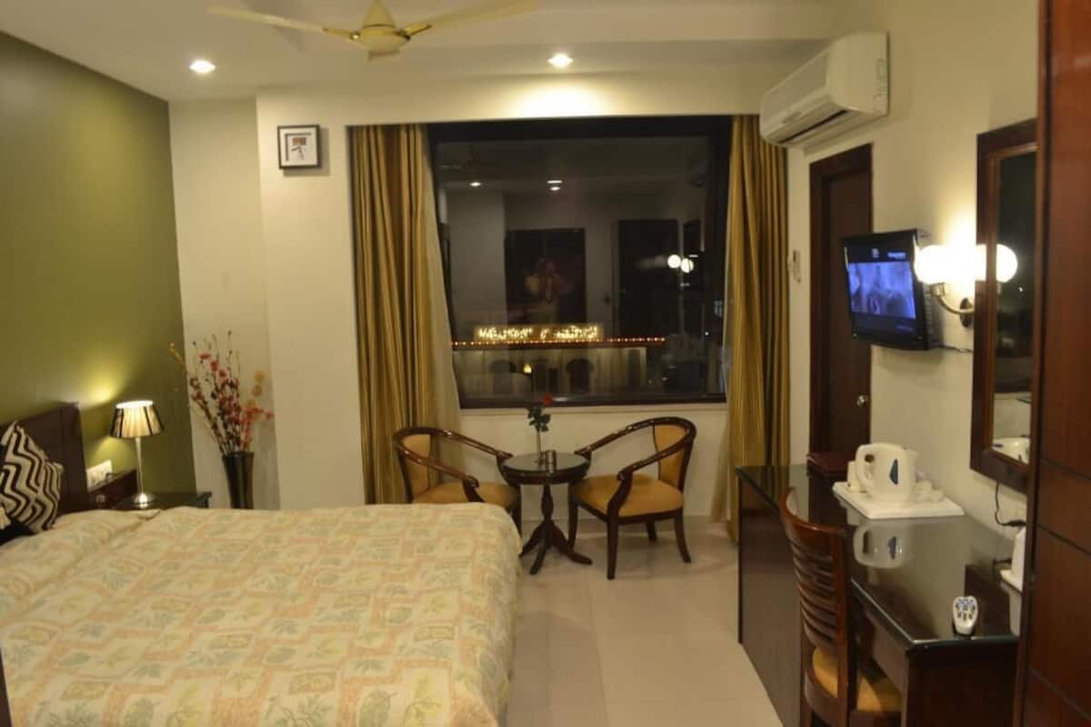 Quarto de hotel com cama, uma mesinha com cadeiras, televisão e janela com cortinas.