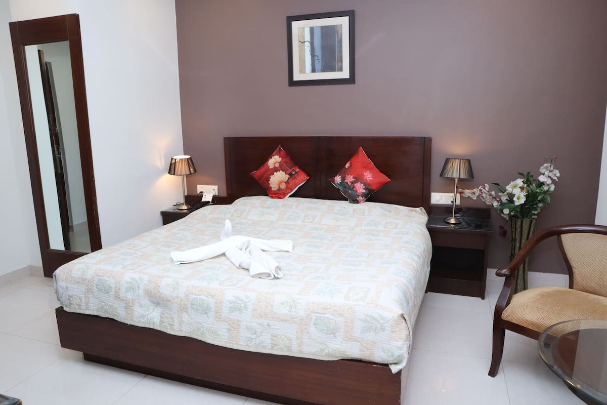 Uma cama bem arrumada com colcha florida, duas almofadas decorativas, uma mesa de cabeceira com luminária, um espelho e uma cadeira.