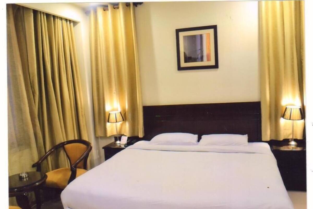 Quarto de hotel com uma cama grande, duas luminárias de cabeceira, uma cadeira, uma mesinha e cortinas.