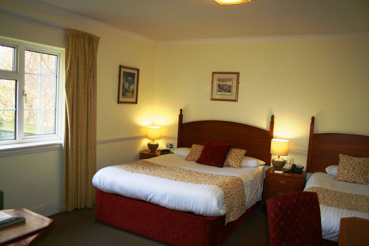 Quarto de hotel com duas camas de solteiro, cabeceira de madeira, mesa de cabeceira com abajur e uma janela com cortinas.