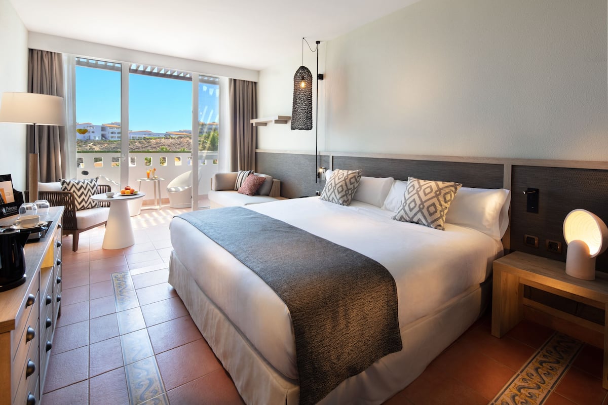 Quarto de hotel com uma cama grande, uma mesa com cafeteira, uma cadeira, uma mesinha e vista para a cidade através de grandes janelas.