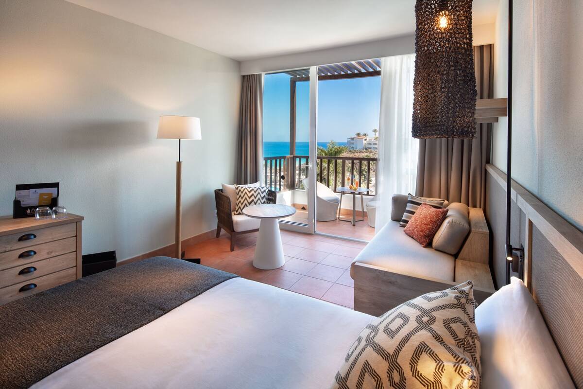 Quarto de hotel com cama, escrivaninha, cadeira, mesa pequena e varanda vista para o mar.