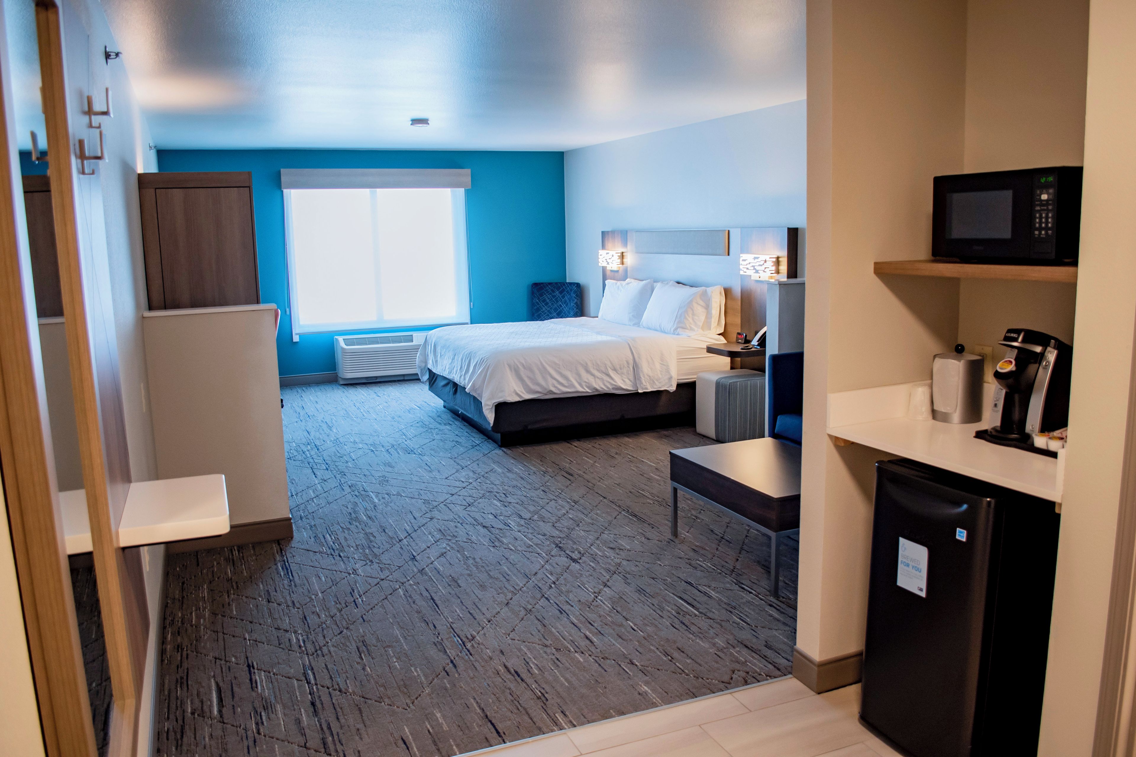 Quarto de hotel com cama, mesa de cabeceira, luminária, micro-ondas e mini-geladeira.