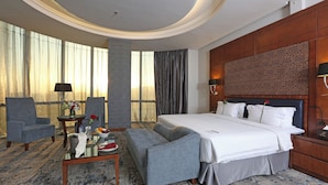 Quarto de hotel com uma cama grande, duas poltronas, uma mesinha com um vaso de flores e vista para o exterior através de janelas amplas.