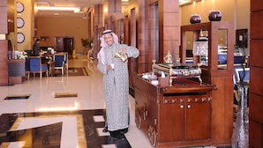 Um homem vestido com traje tradicional segura uma chaleira em um saguão de hotel, com balcão de recepção, cadeiras e relógios nas paredes.