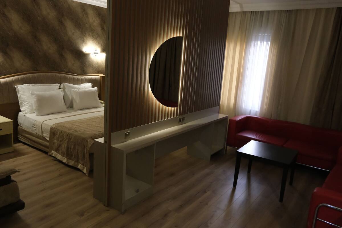 Quarto de hotel com cama, mesa de cabeceira, sofá vermelho, pequena mesa e espelho.