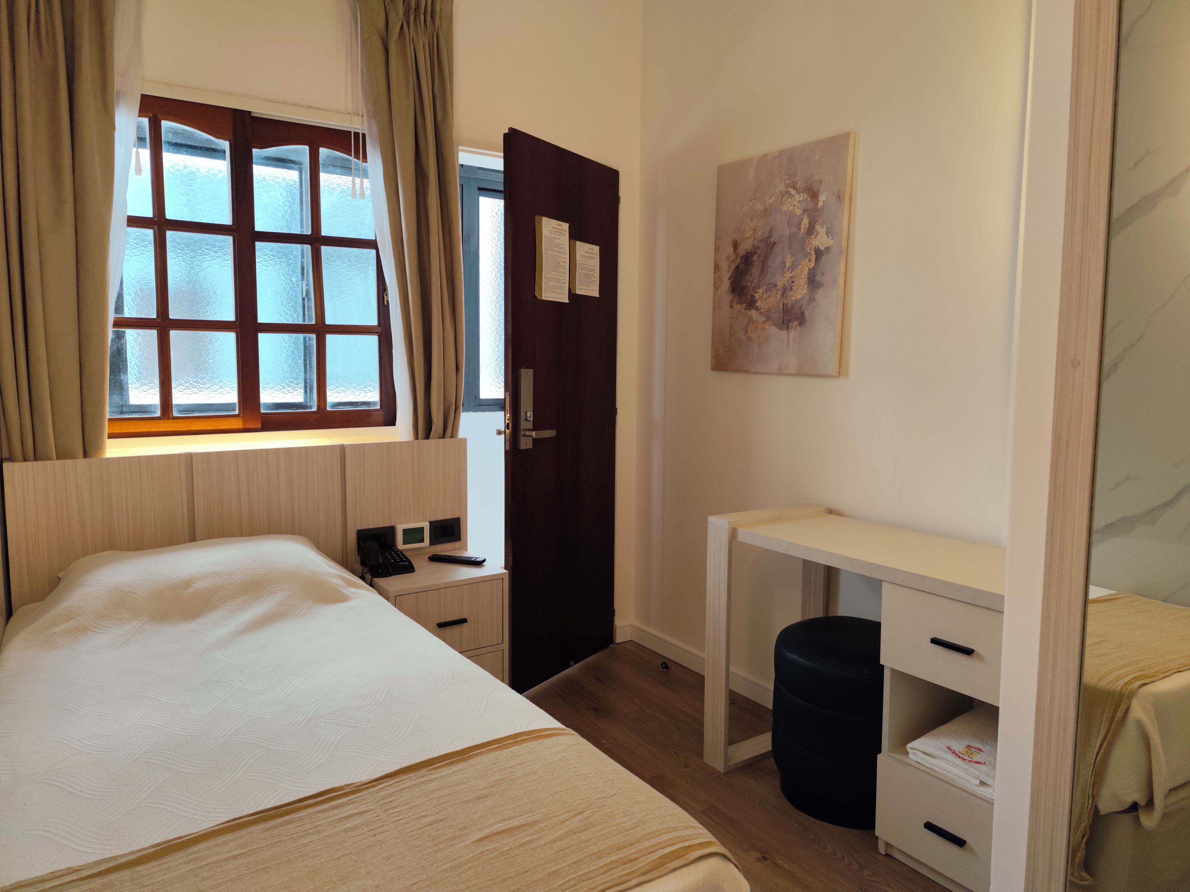 Quarto de hotel com cama, escrivaninha, cadeira, lixeira, porta e janela com cortinas.
