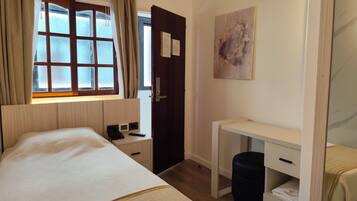 Quarto de hotel com cama, escrivaninha, cadeira, lixeira, porta e janela com cortinas.